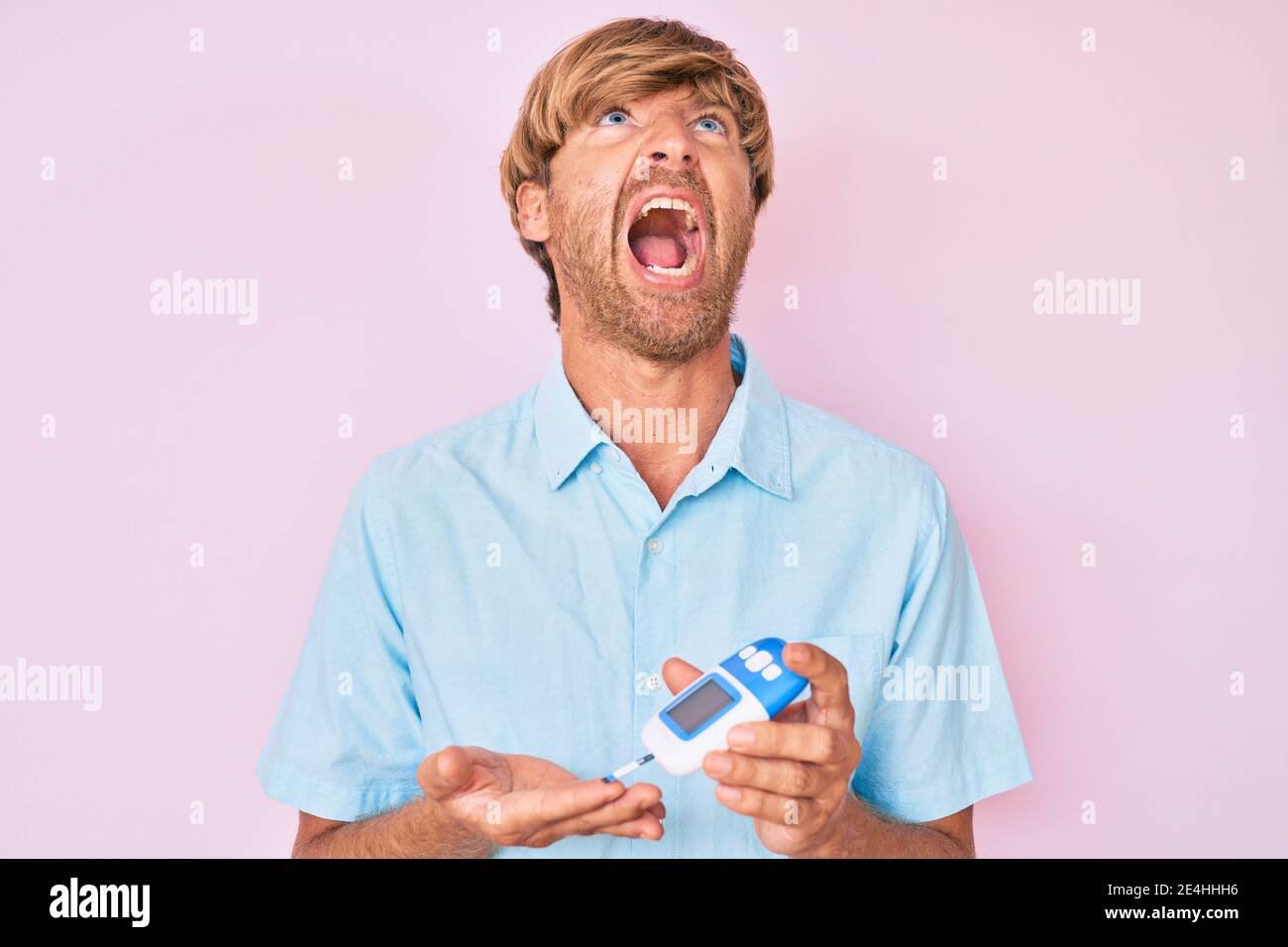 Young blond man using glucose meter angry and mad screaming frustrated ...