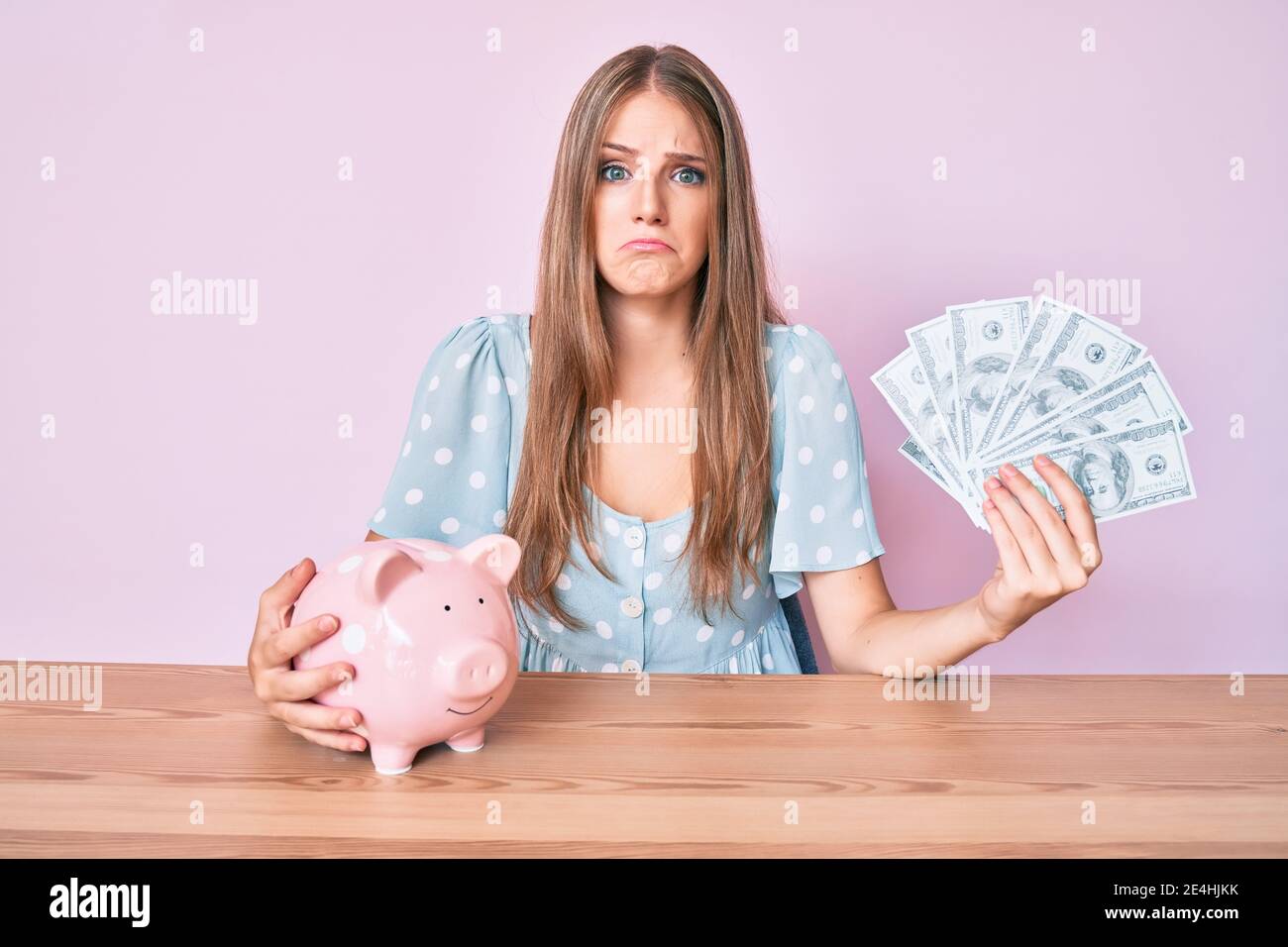 Young blonde girl holding dollars and piggy bank sitting on the table ...