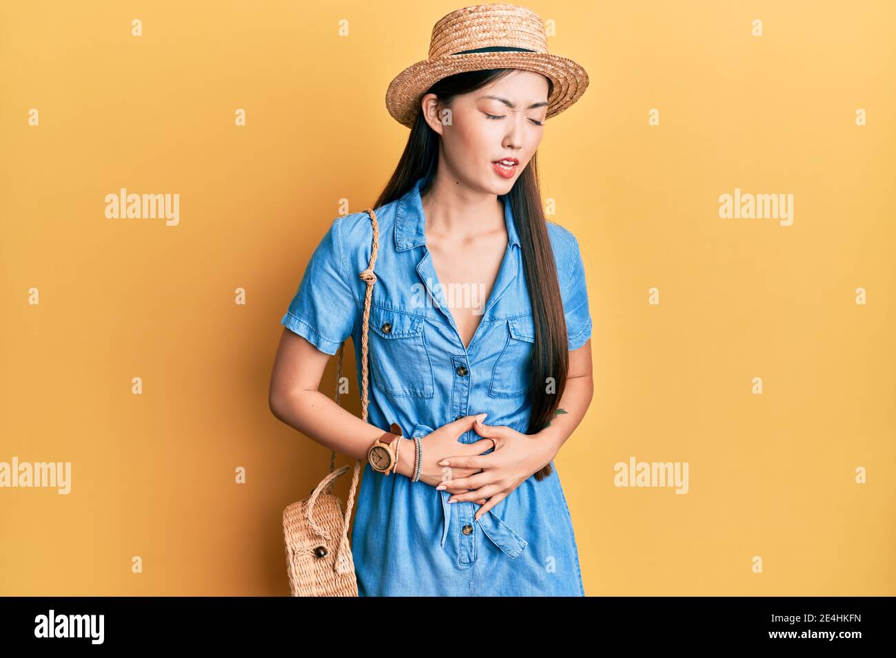 Young chinese woman wearing summer hat with hand on stomach because ...