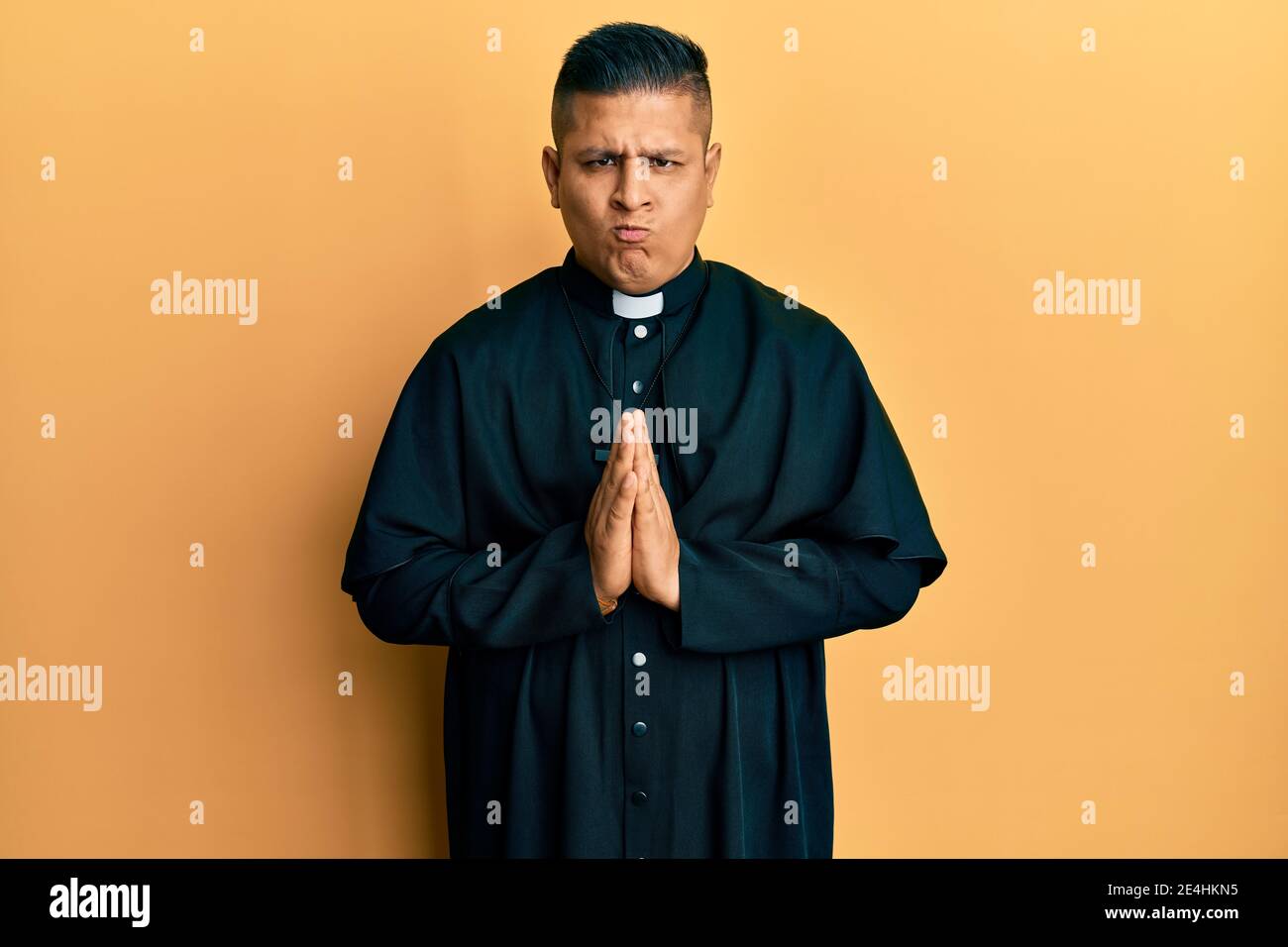 Young latin priest man praying with hands together skeptic and nervous ...