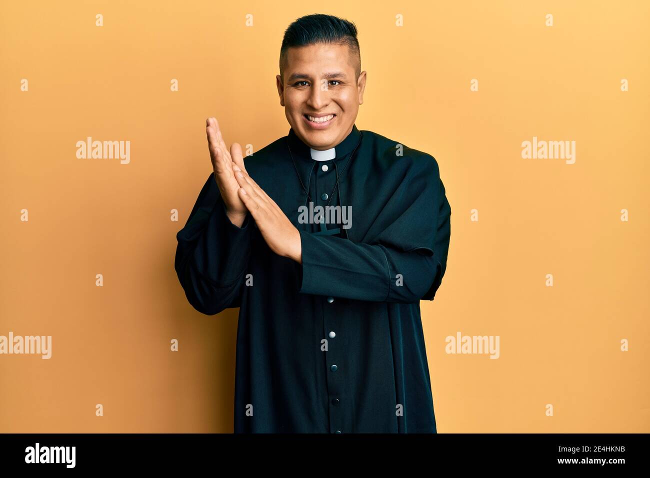 Young latin priest man standing over yellow background clapping and ...