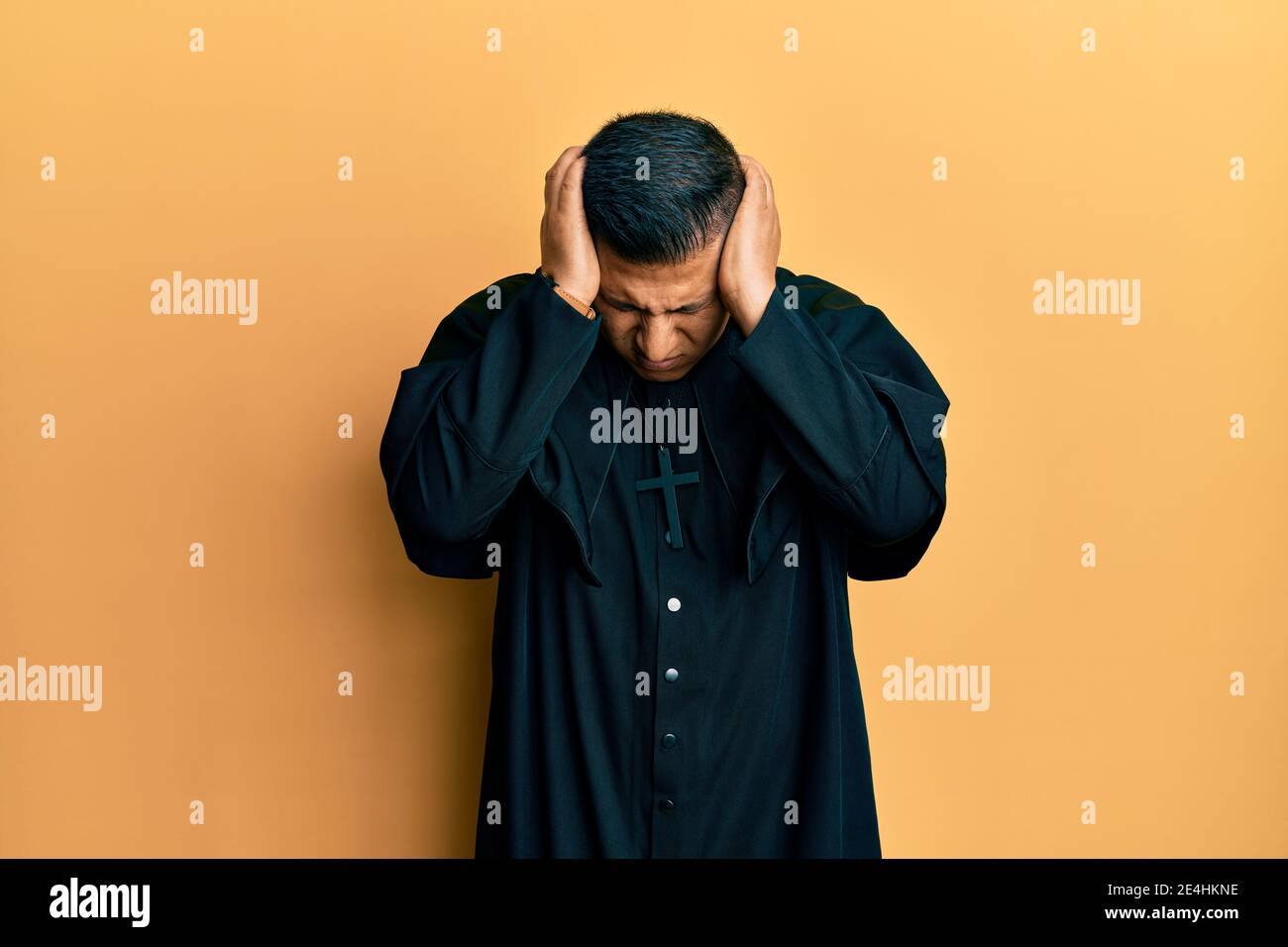 Young latin priest man standing over yellow background suffering from ...