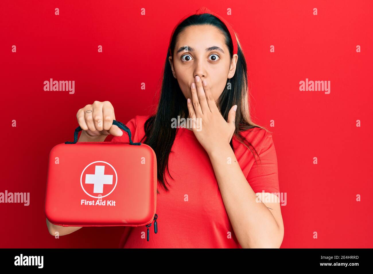 Young hispanic woman holding first aid kit covering mouth with hand ...