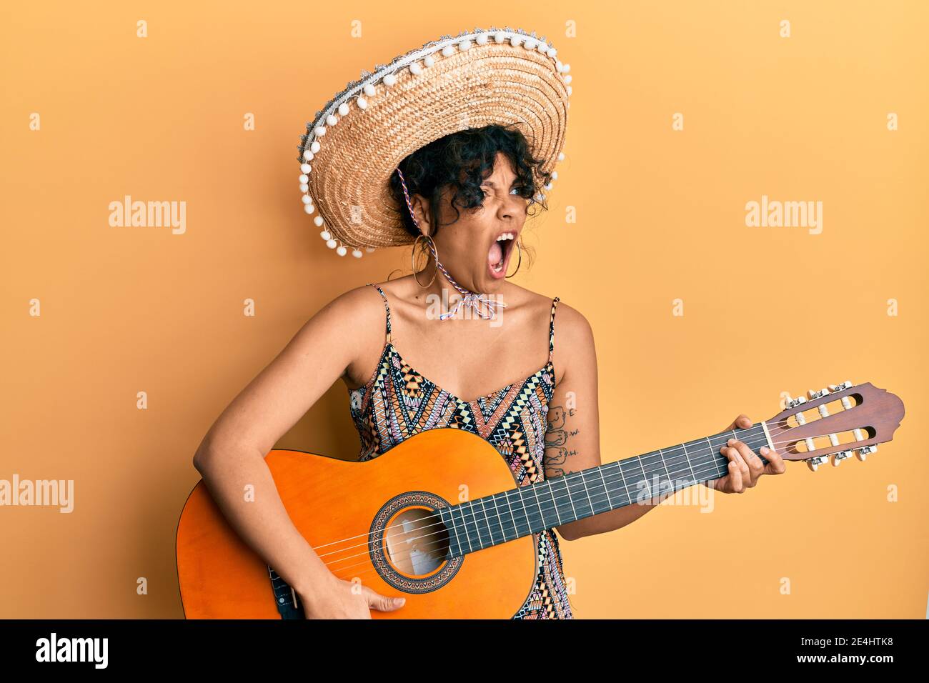 Young hispanic woman holding mexican hat playing classical guitar angry ...