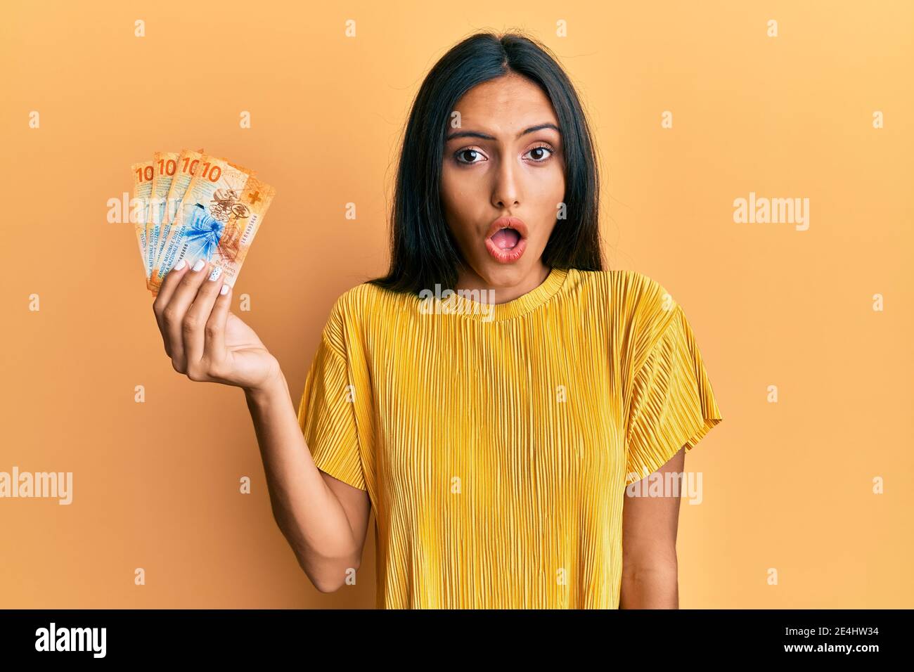 Young brunette woman holding 10 swiss franc banknotes scared and amazed ...