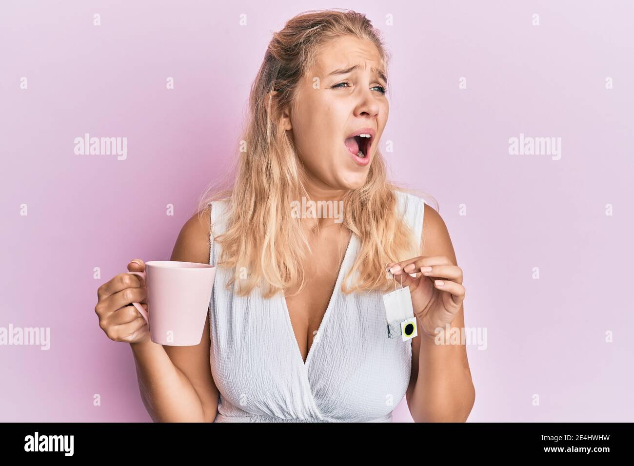 Young blonde girl holding cup of tea angry and mad screaming frustrated ...