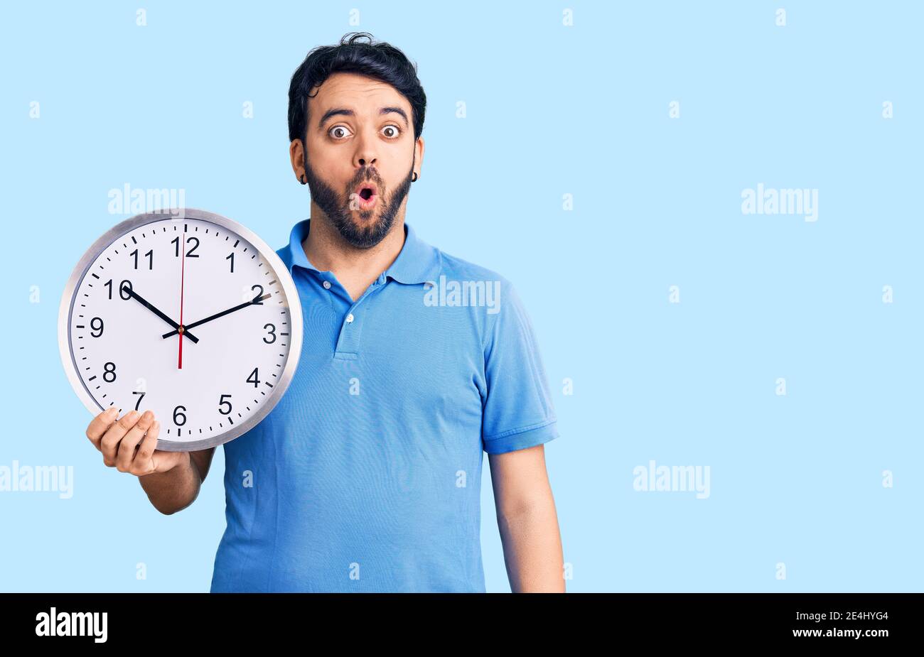 Young hispanic man holding big clock scared and amazed with open mouth ...