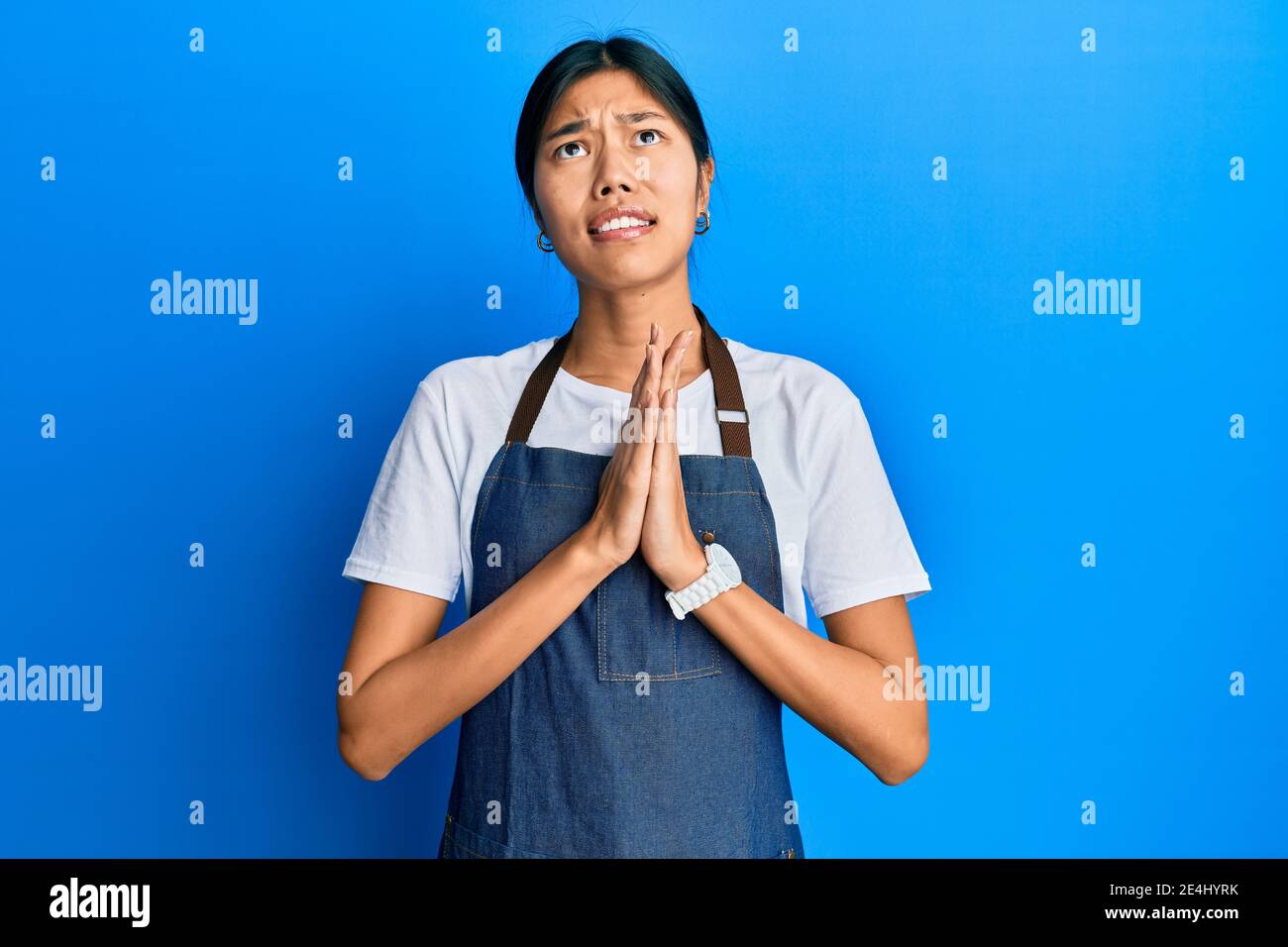 Young chinese woman wearing waiter apron begging and praying with hands ...