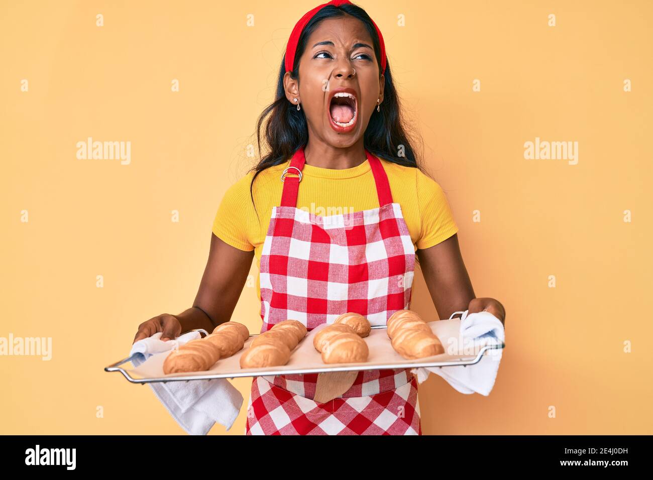 Young indian girl wearing baker uniform holding homemade bread angry ...