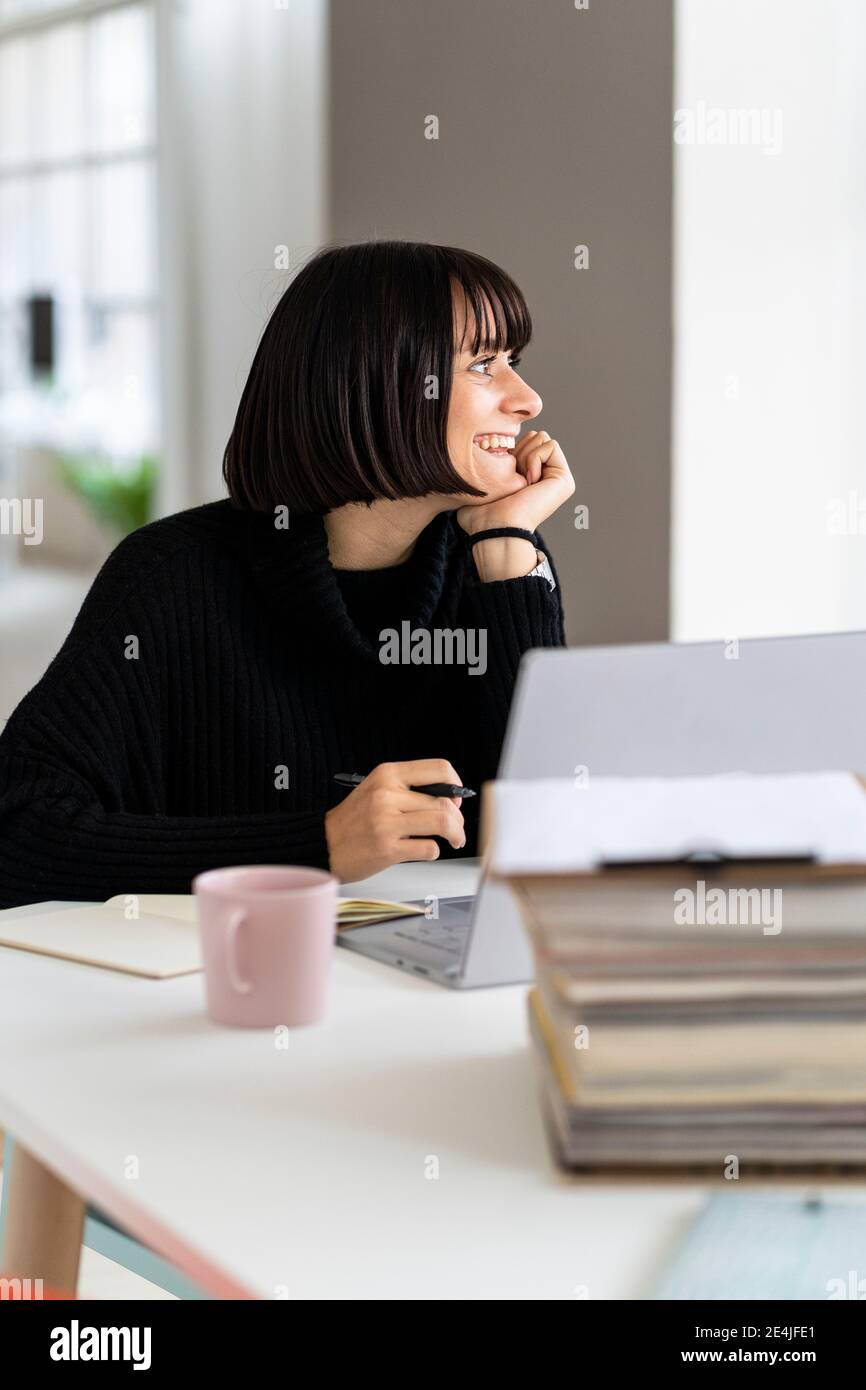 Smiling young female student day dreaming while studying in study room ...