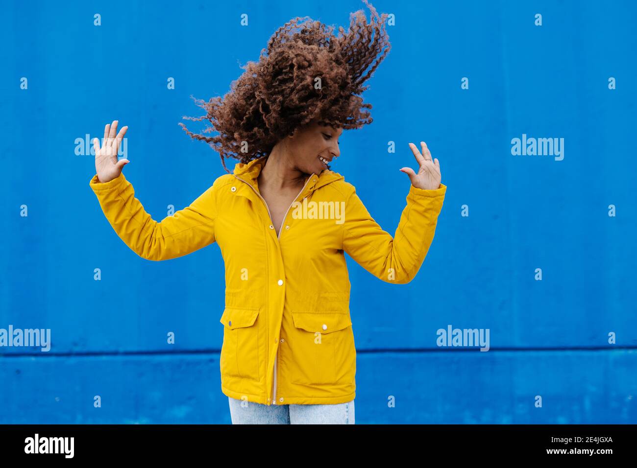 Playful woman tossing hair while standing against blue wall Stock Photo ...