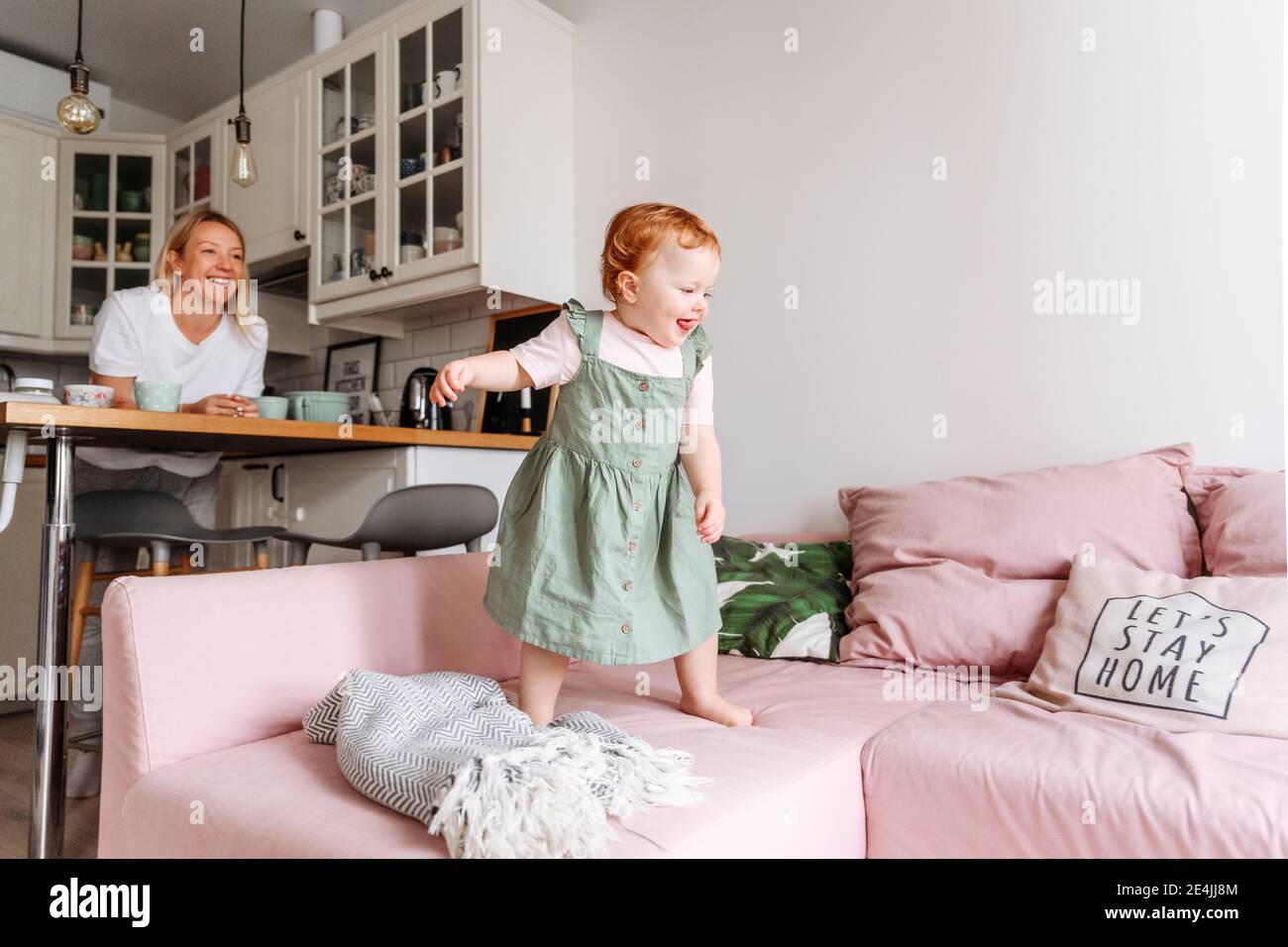 Baby girl jumping on sofa, smiling mother in background Stock Photo - Alamy