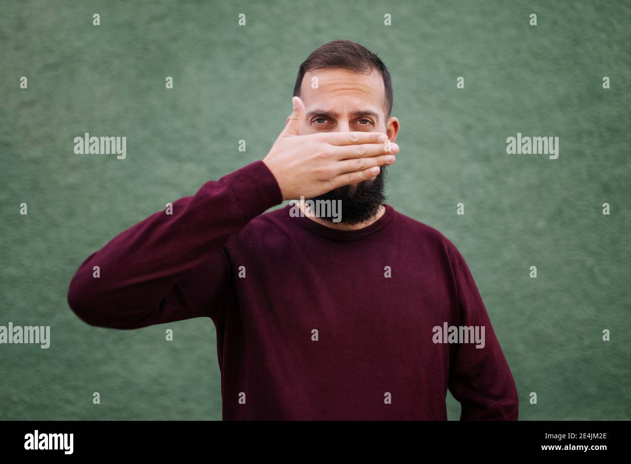 Mid adult man hiding face with hand while standing against green wall ...