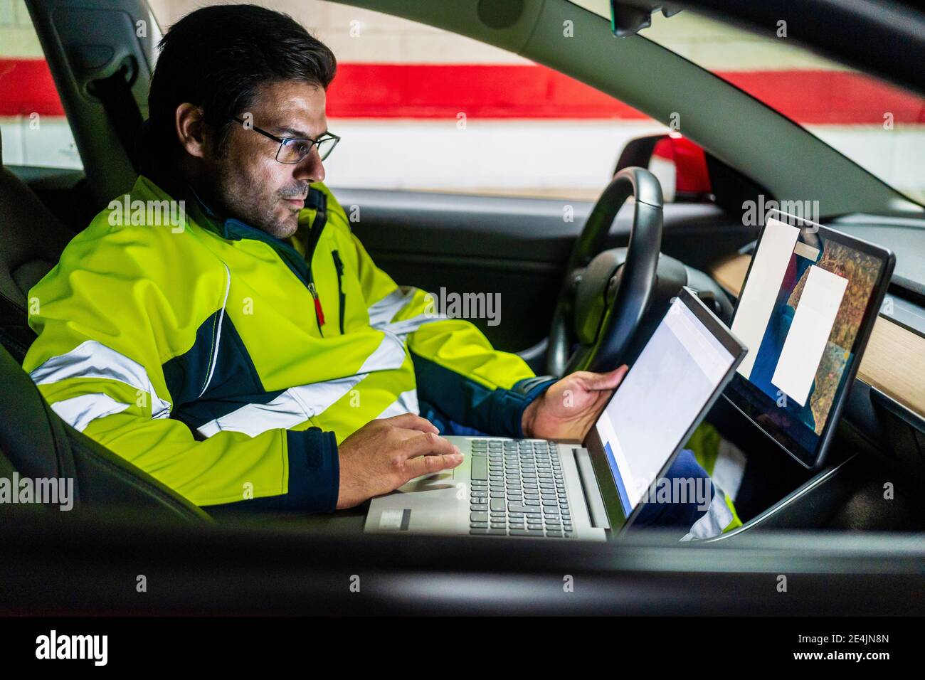 Mature technician programming on laptop while sitting in electric car ...