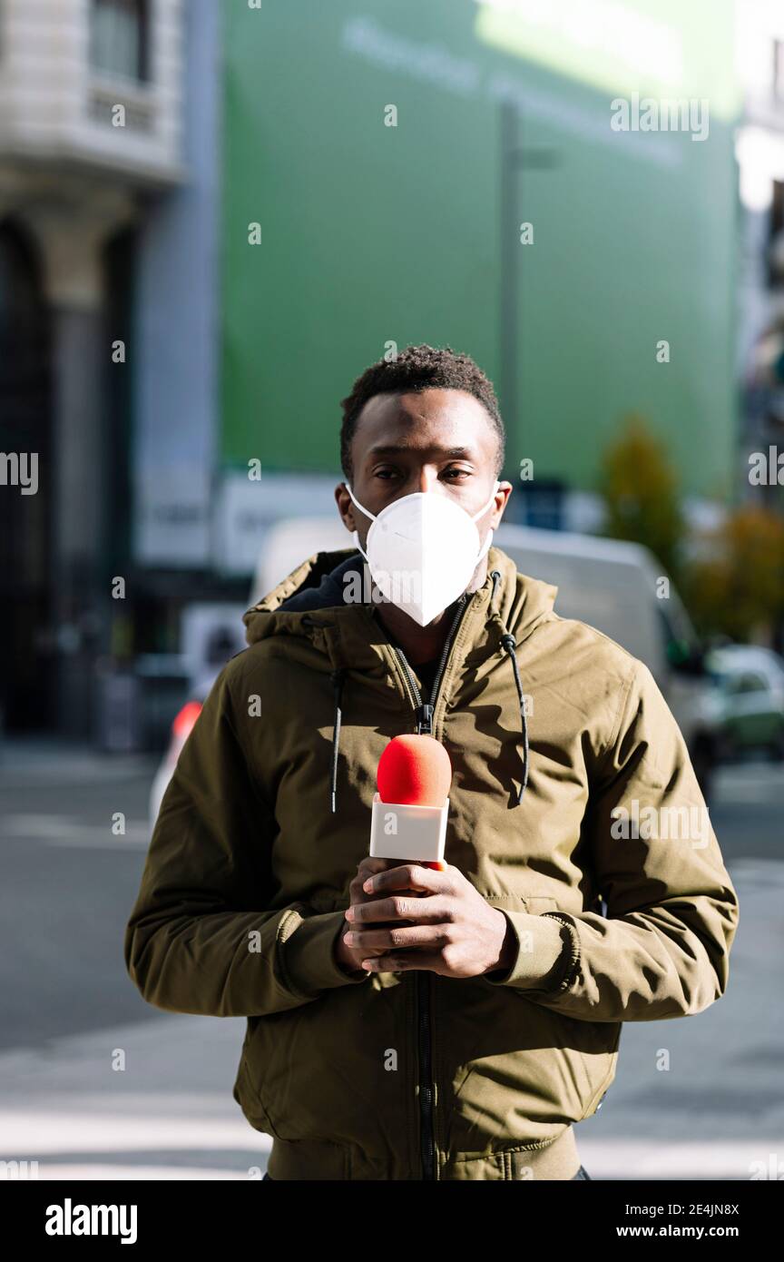 Male TV reporter wearing face mask holding microphone while standing on ...
