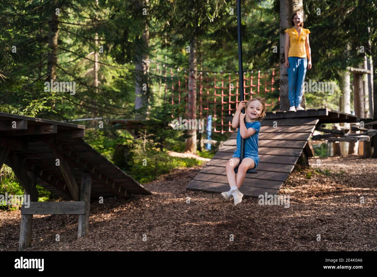 Little girl riding forest zip line with mother standing in background ...