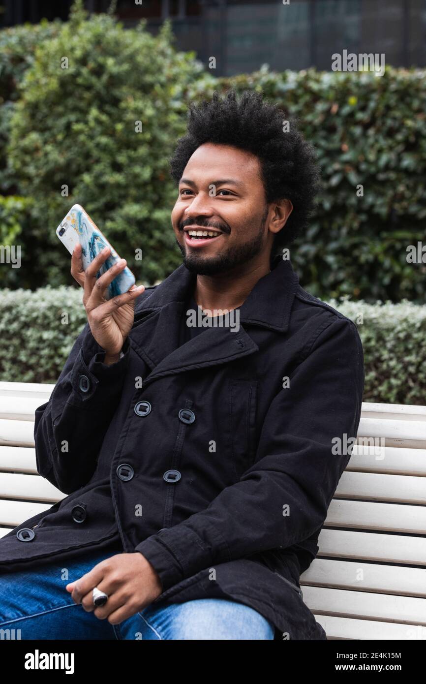 Happy man wearing coat talking over mobile phone while sitting on bench ...
