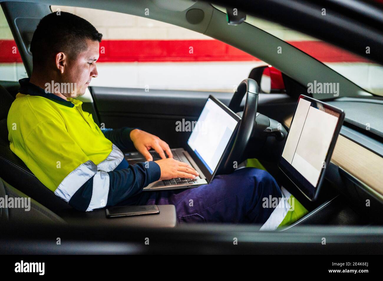 Male technician programming on laptop while sitting in electric car ...