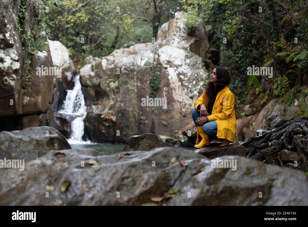 Thoughtful female hiker crouching on rock against waterfall in forest ...
