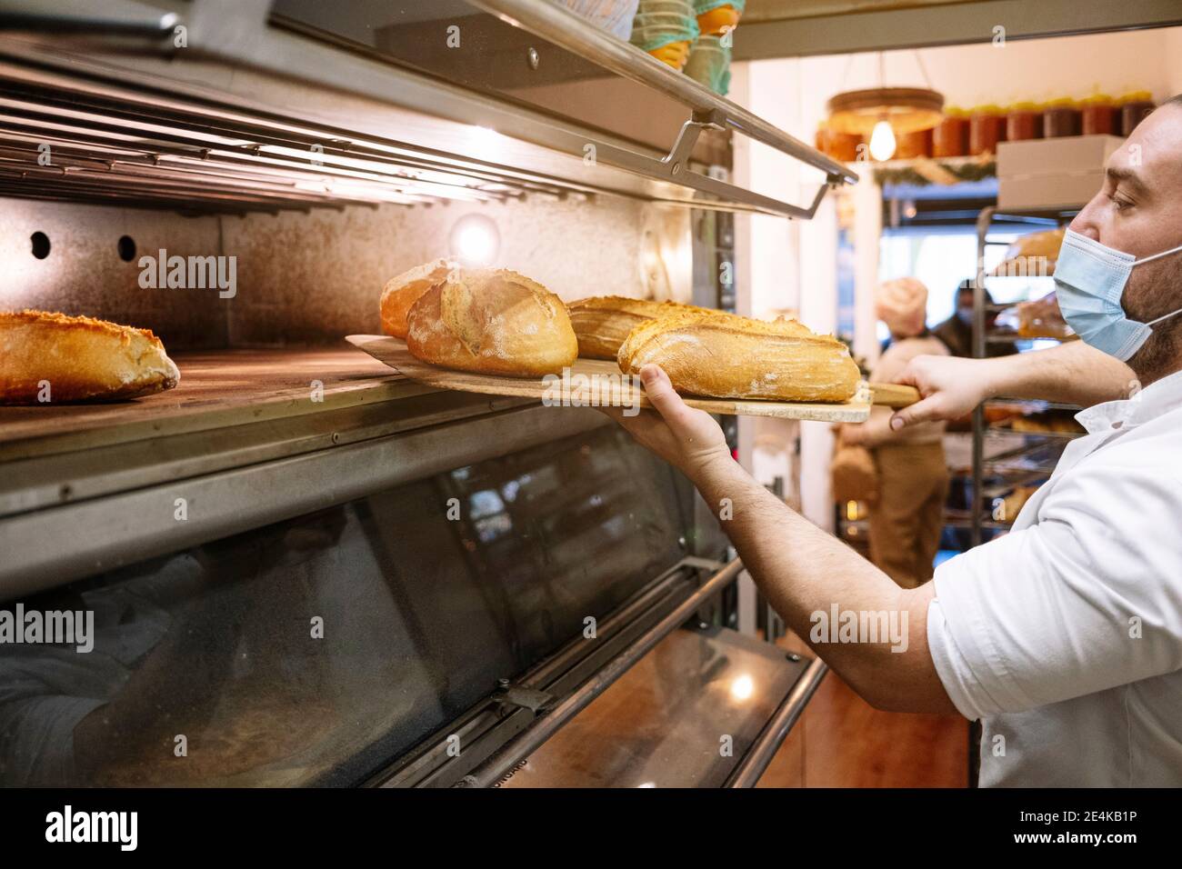Male chef in protective face mask removing fresh bread from oven in ...