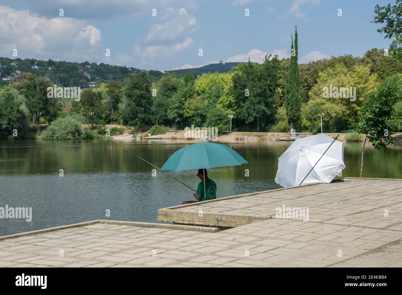 Man with an umbrella fishing in the public park pond, peaceful lake ...