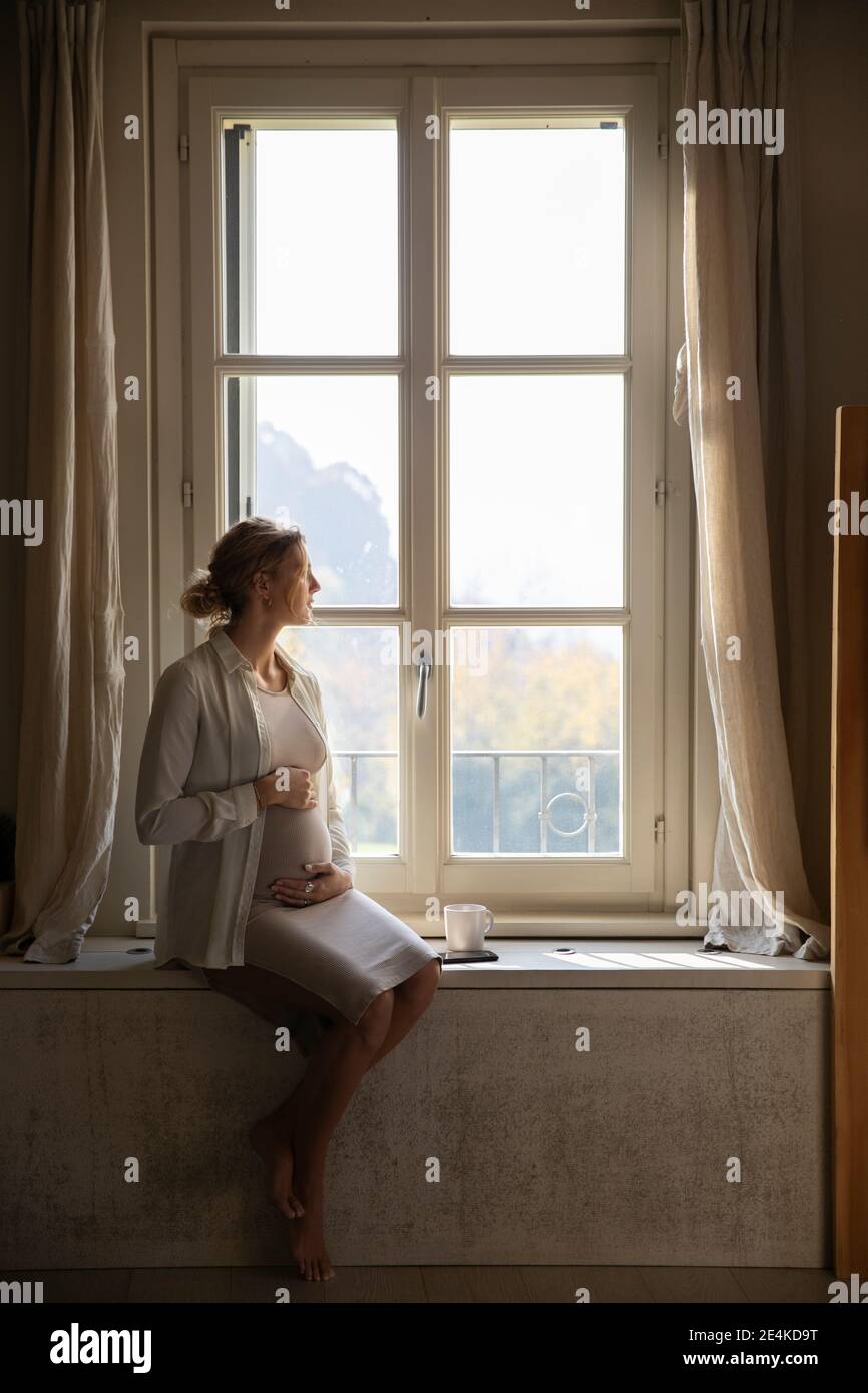 Pregnant woman with hands on stomach looking through window at home ...