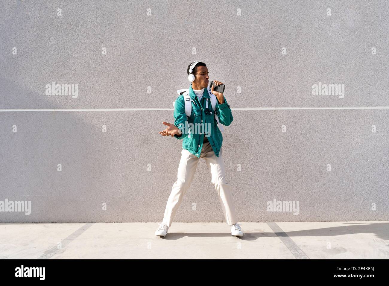 Young man with headphones and backpack singing while standing against ...