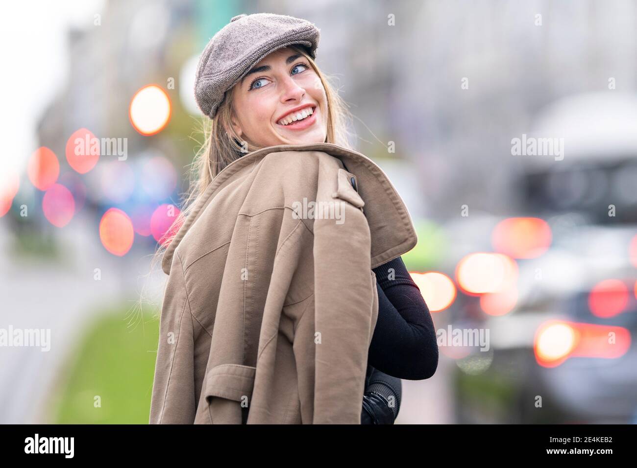 Happy woman holding overcoat looking over shoulder while standing on ...