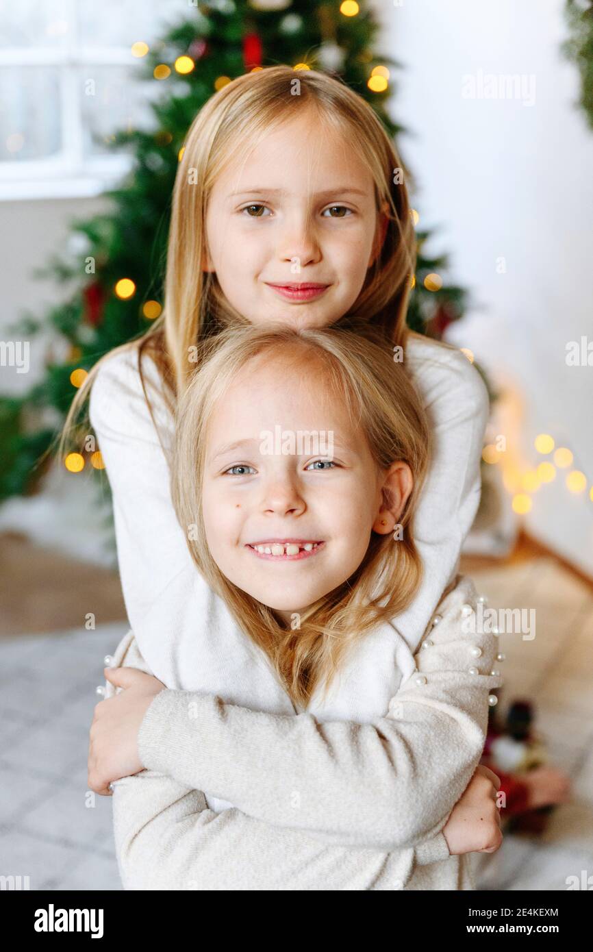 Smiling sisters embracing with Christmas tree in background at home ...