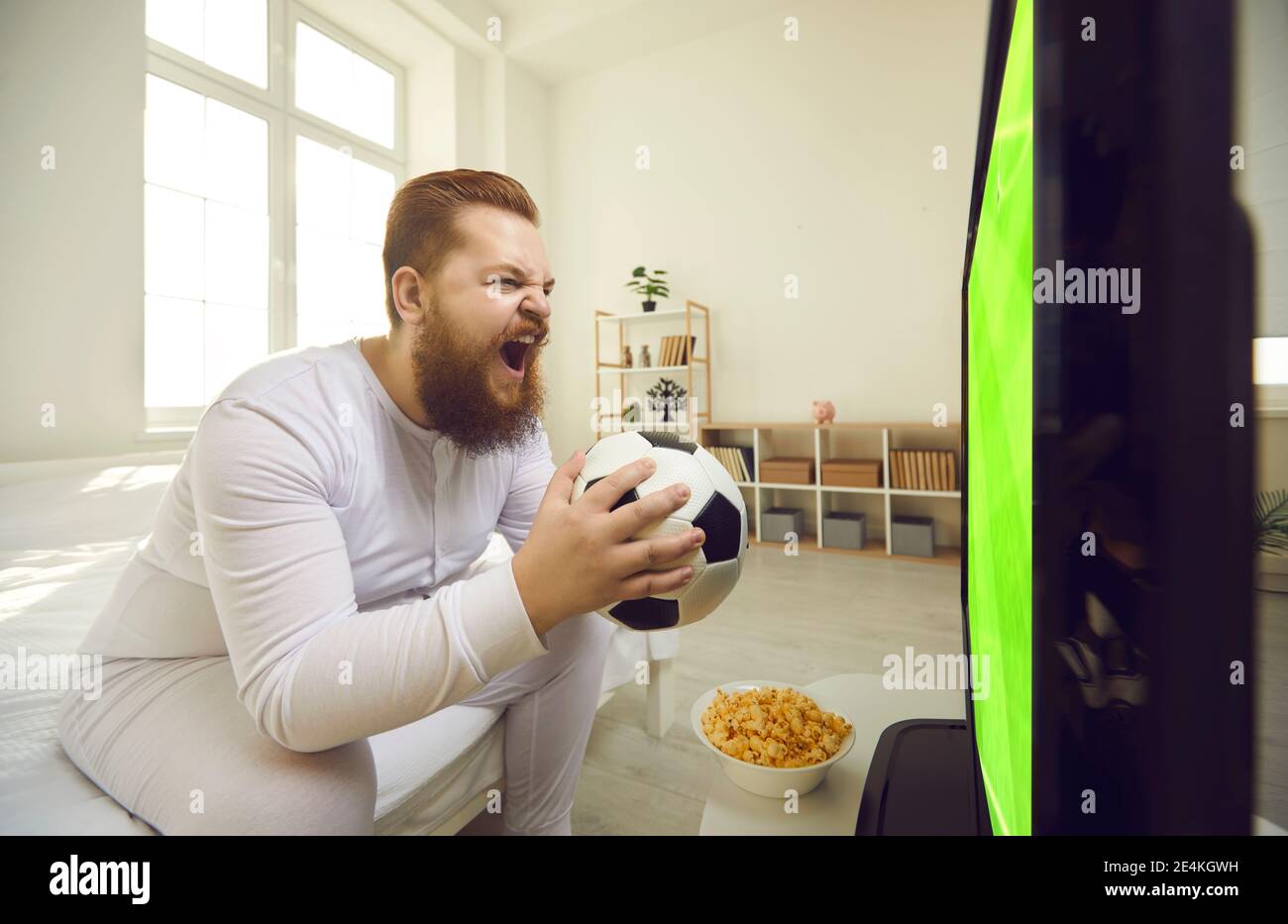 Funny chubby fan sitting on bed in pajamas and watching exciting soccer ...