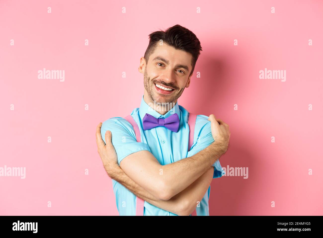 Cheerful young man smiling and hugging himself, wrap hands around own ...
