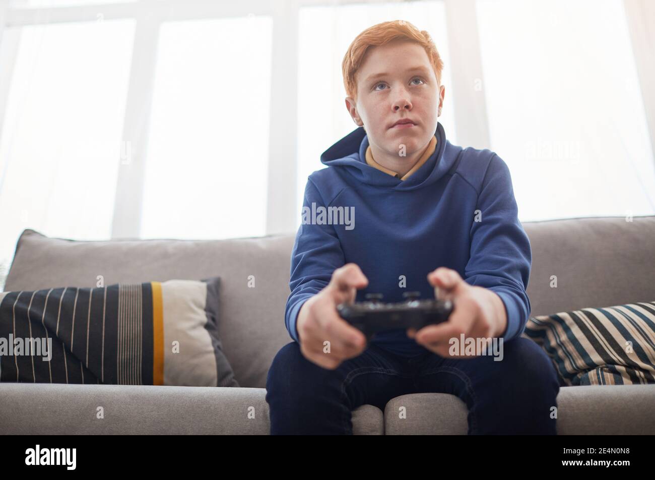 Low angle portrait of red haired teenage boy playing video games while ...