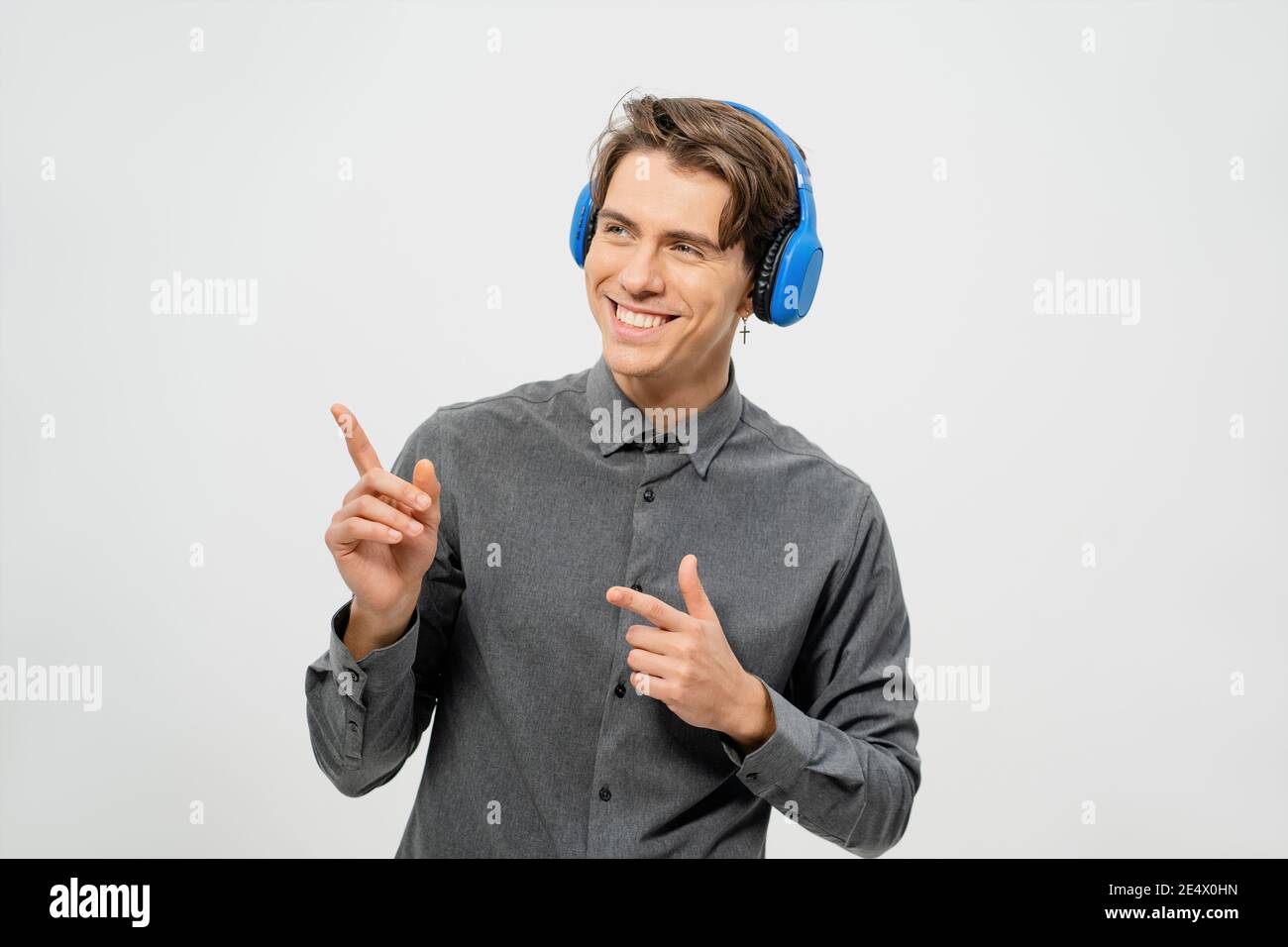 Happy smiling young guy in grey shirt standing listening music wearing ...