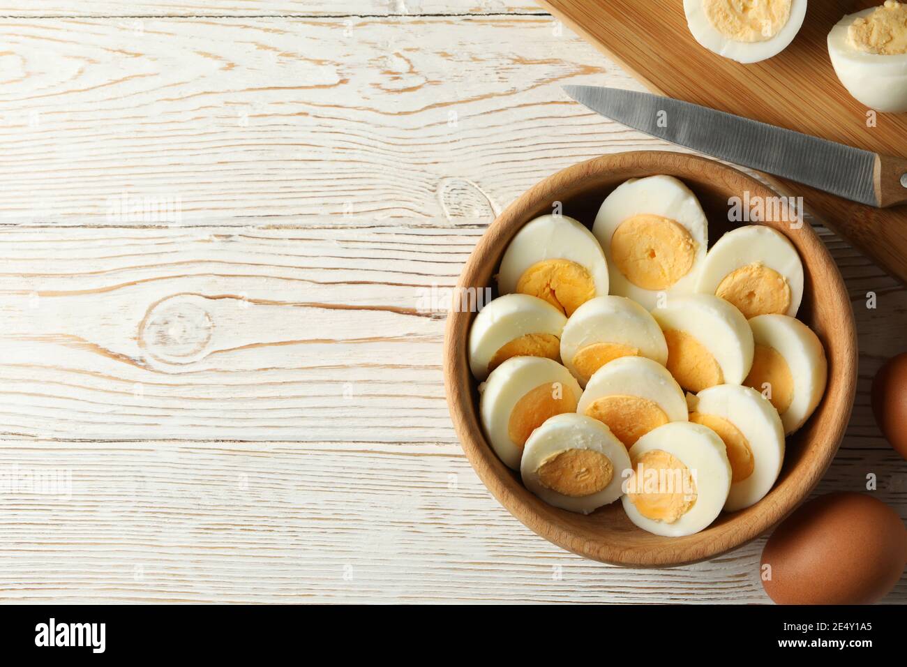 Board and bowl with boiled eggs and knife on white wooden background ...