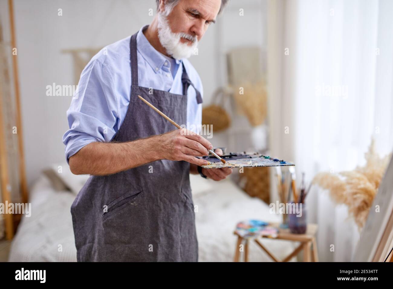 artist man is drawing on easel canvas, senior gray haired man in apron ...