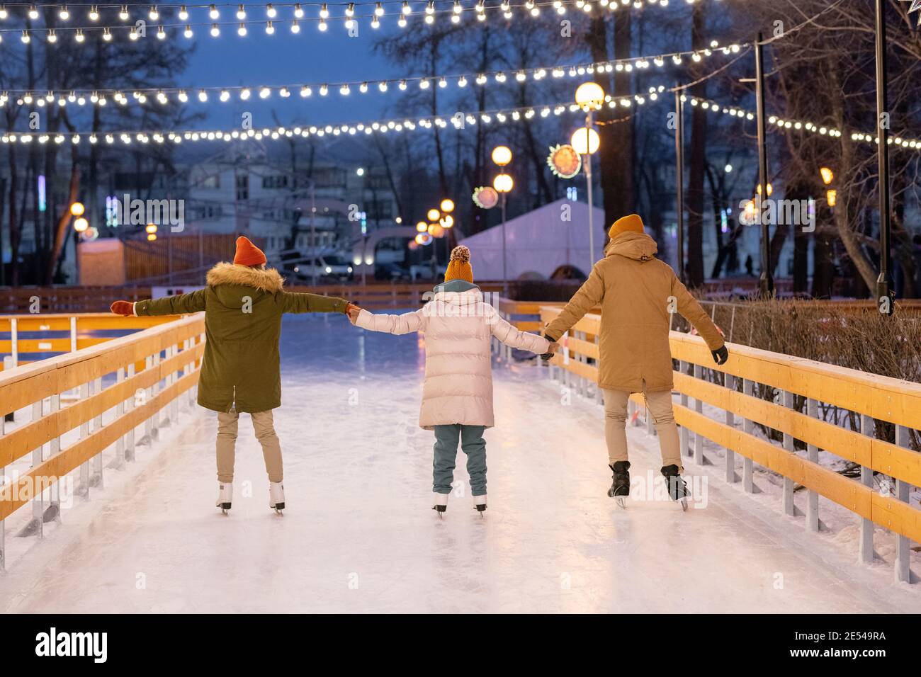 Rear view of family of three holding hands skating on ice rink together ...