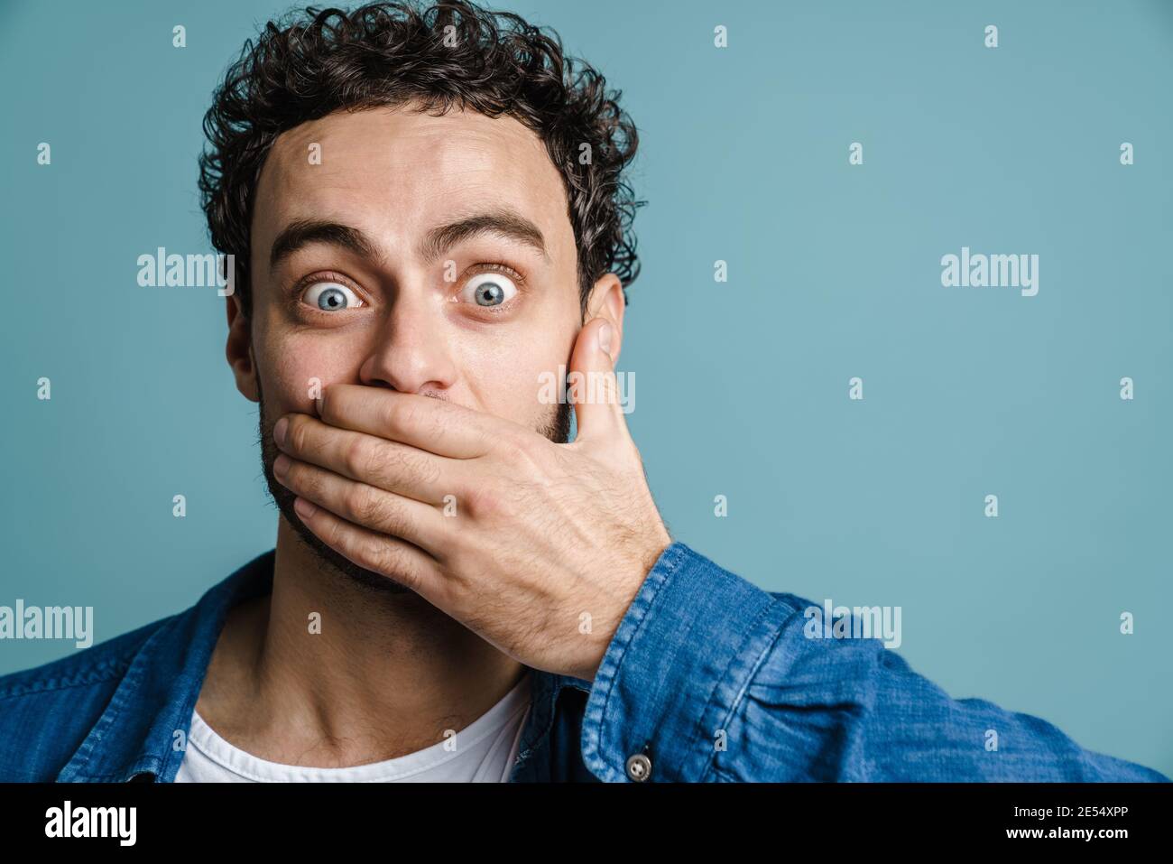Caucasian shocked guy posing and covering his mouth isolated over blue ...