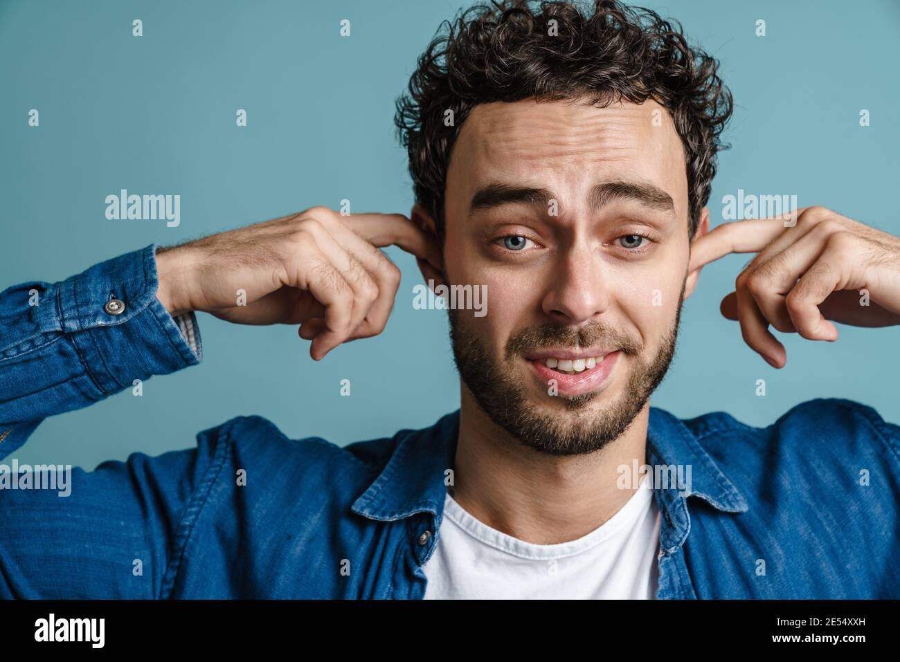 Confused handsome guy posing and plugging his ears isolated over blue ...
