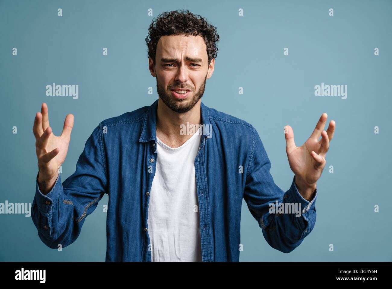 Unhappy handsome guy posing with hands up on camera isolated over blue ...