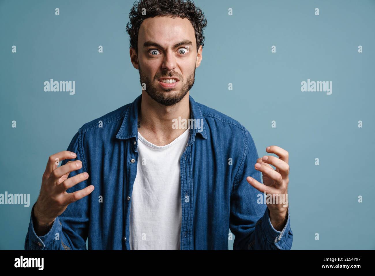 Furious handsome guy posing with hands up on camera isolated over blue ...