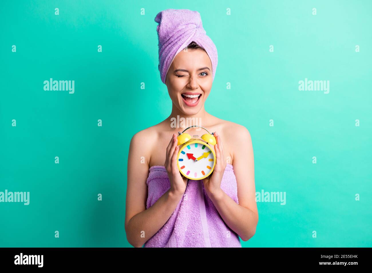Photo of charming lady blink eye hold alarm clock procedure time wear purple towels isolated ...