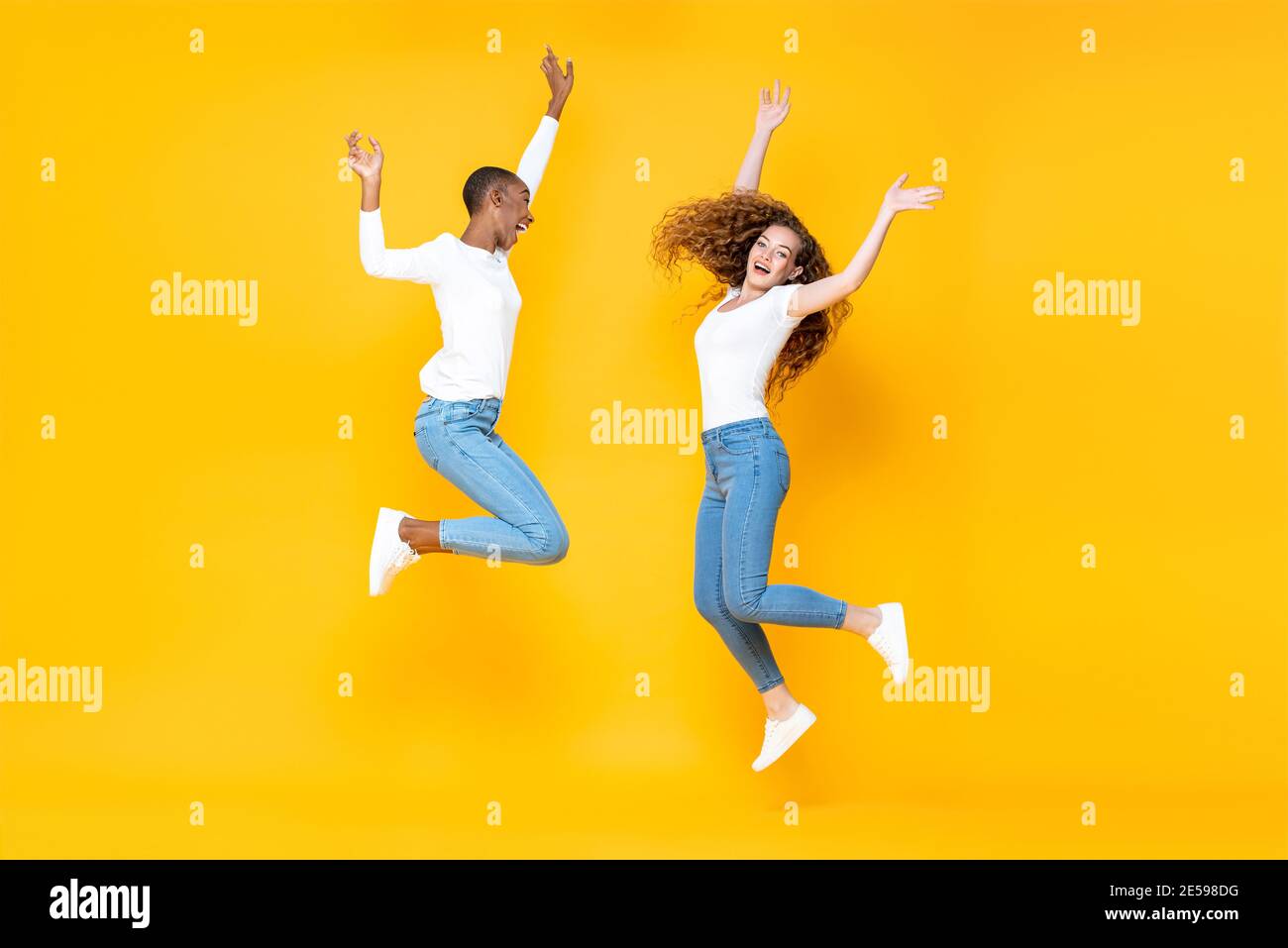 Two interracial woman friends jumping freely in isolated studio yellow ...