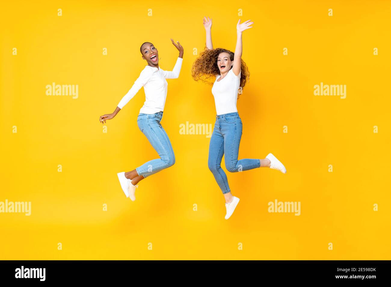 Two interracial woman friends jumping freely in isolated studio yellow ...