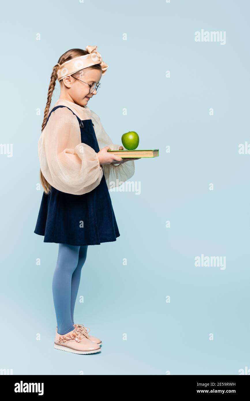 full length of smart kid in glasses looking at book and green apple on ...
