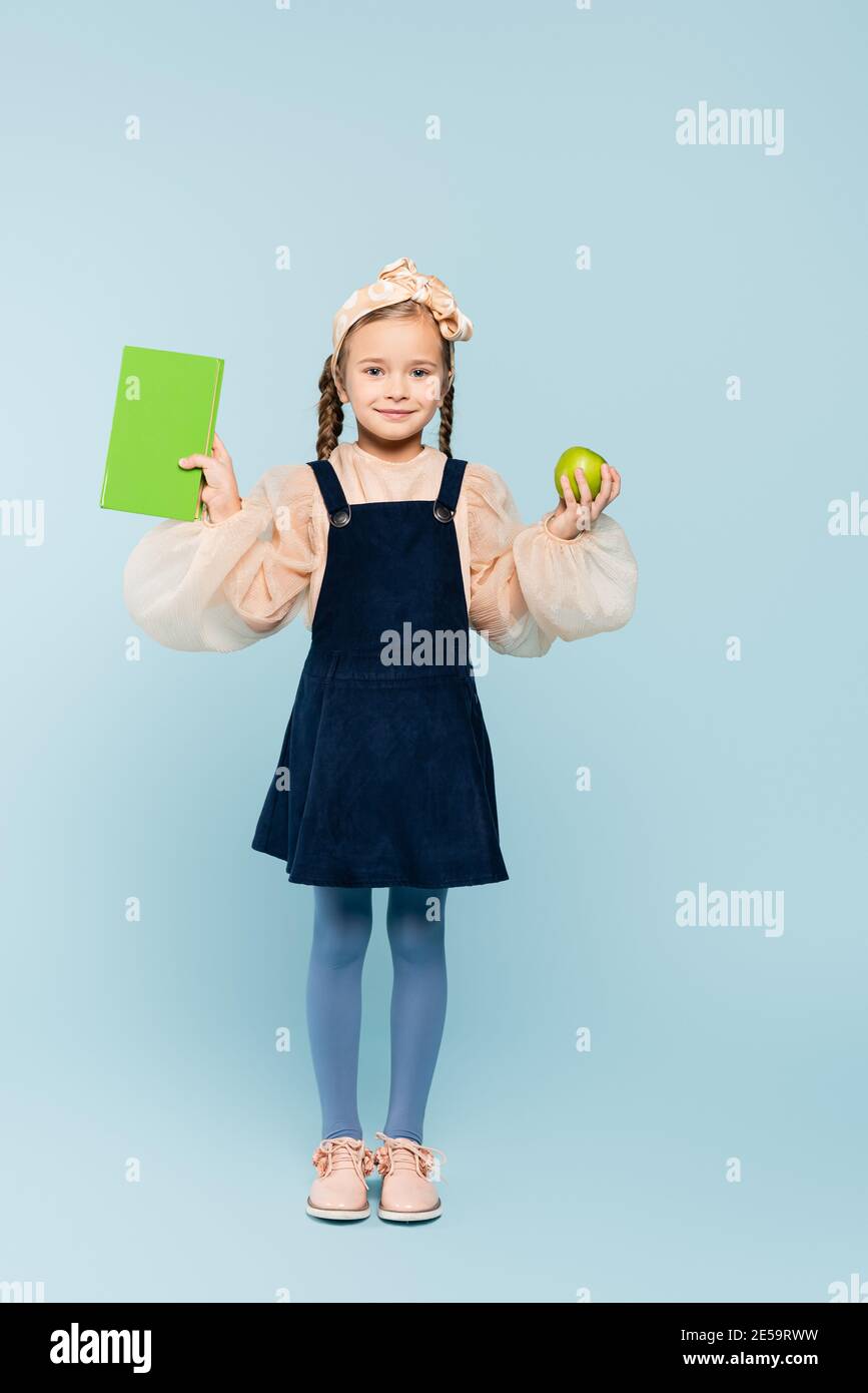 full length of happy kid in dress holding book and green apple on blue ...