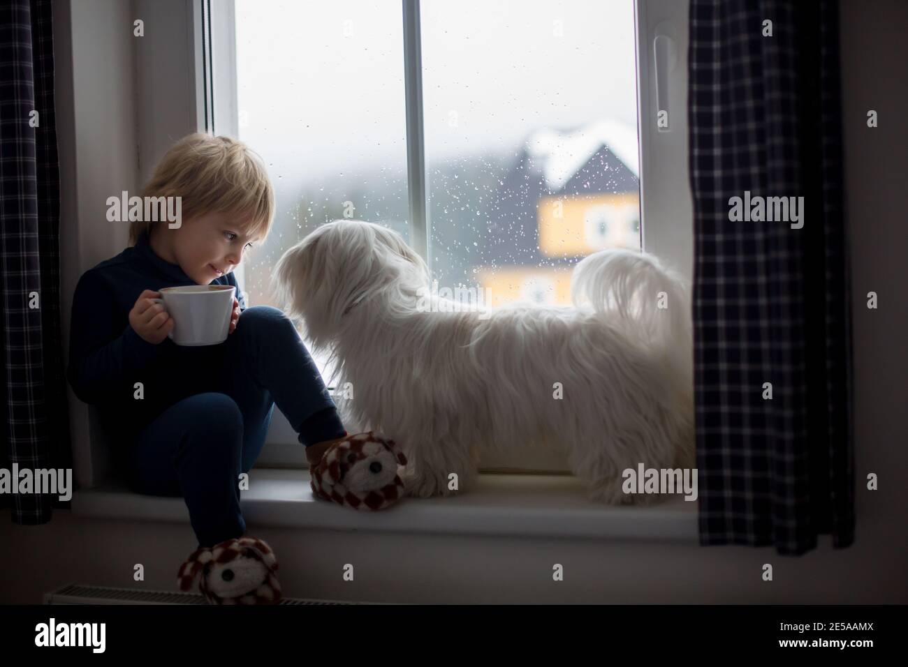 Toddler child, sitting on the window, watching the snow falling ...