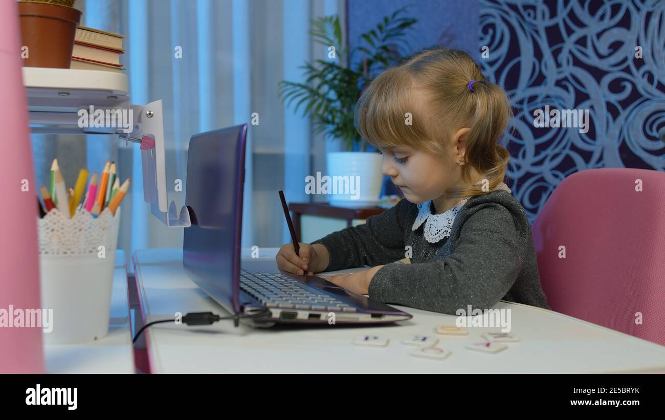 Child girl doing school homework, writes in notebook, using laptop ...