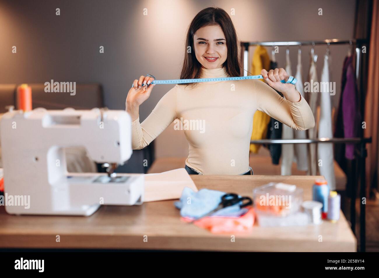 Young dressmaker woman sews clothes on sewing machine. Holding tape ...