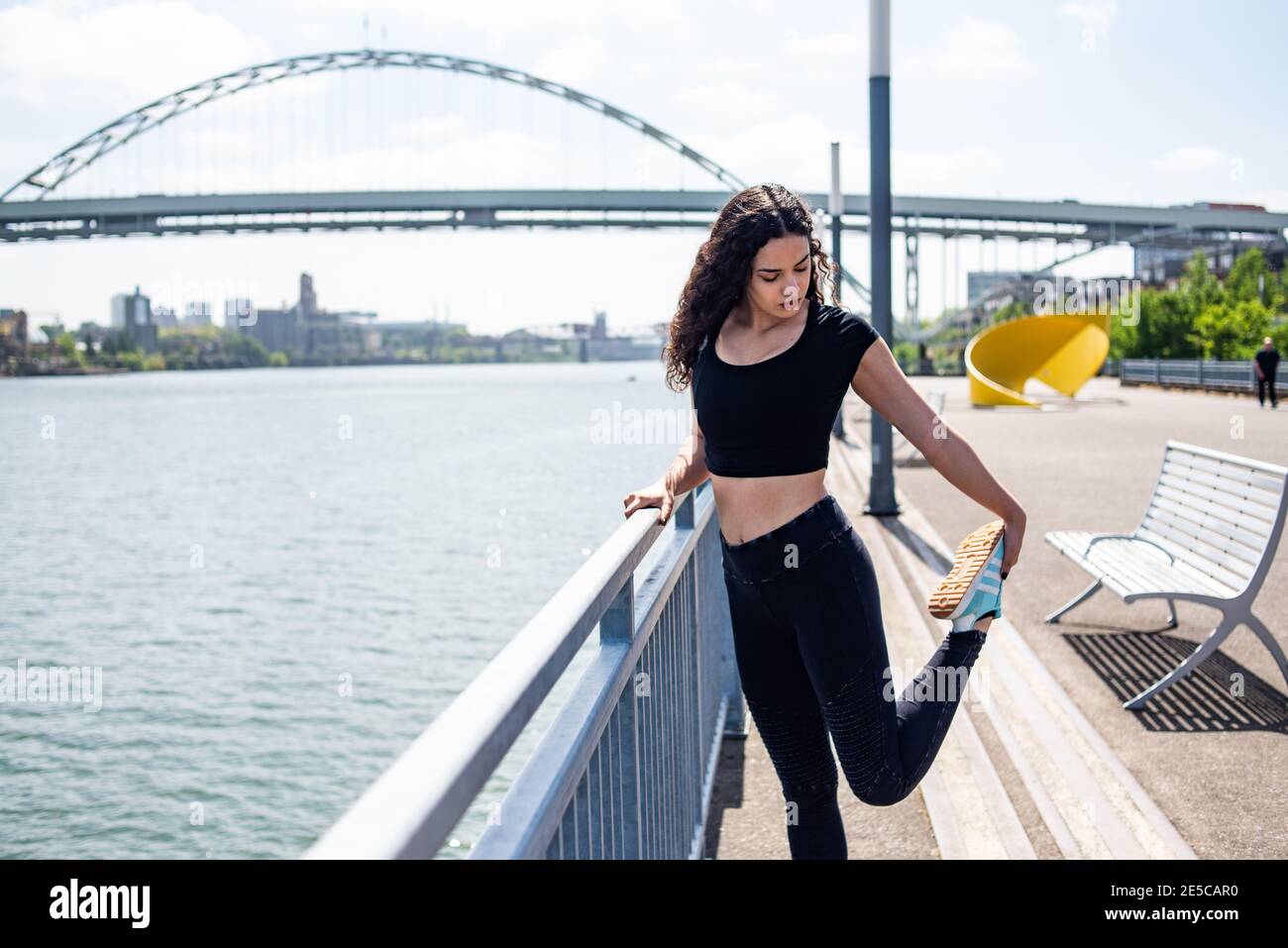 Woman stretching hamstring along riverfront with bridge in background ...