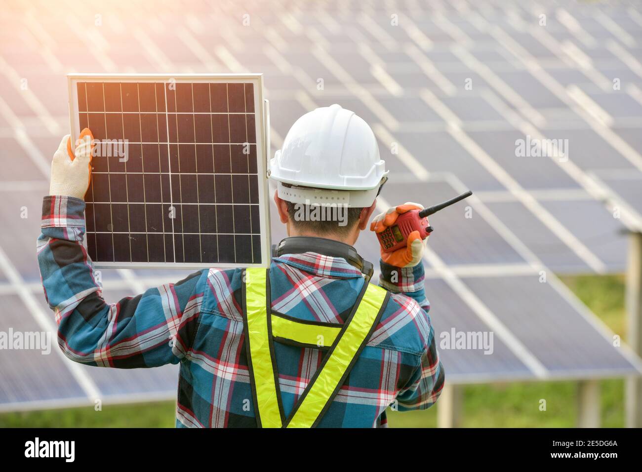 Rear view of an engineer at a solar farm carrying a solar panel ...